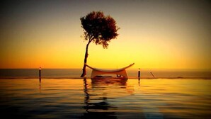 Uma rede amarrada entre dois postes sobre águas calmas, com a silhueta de uma árvore ao pôr do sol.