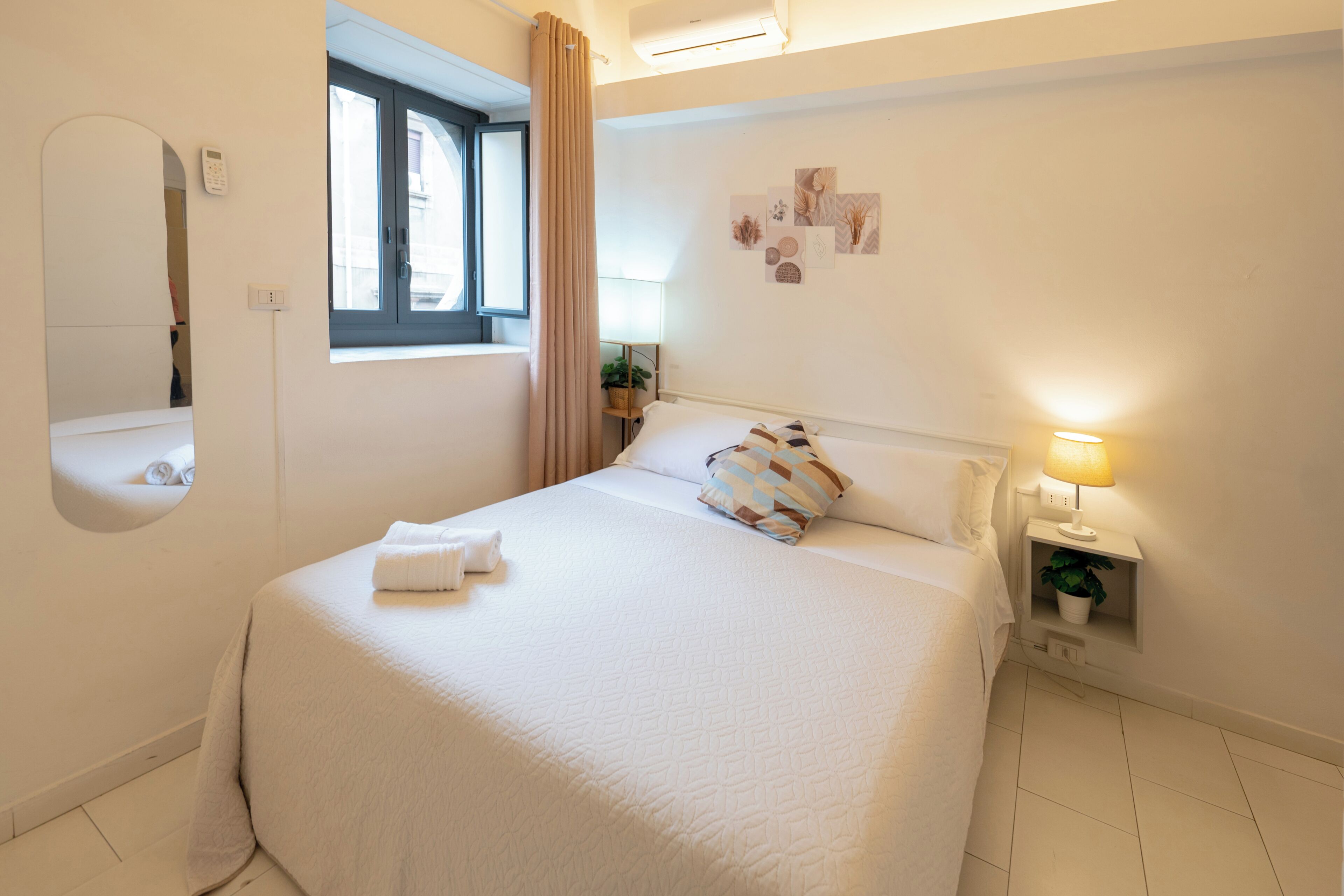 Cama bem arrumada com edredom branco e almofada decorativa, mesa de cabeceira com abajur, e janela com cortinas.