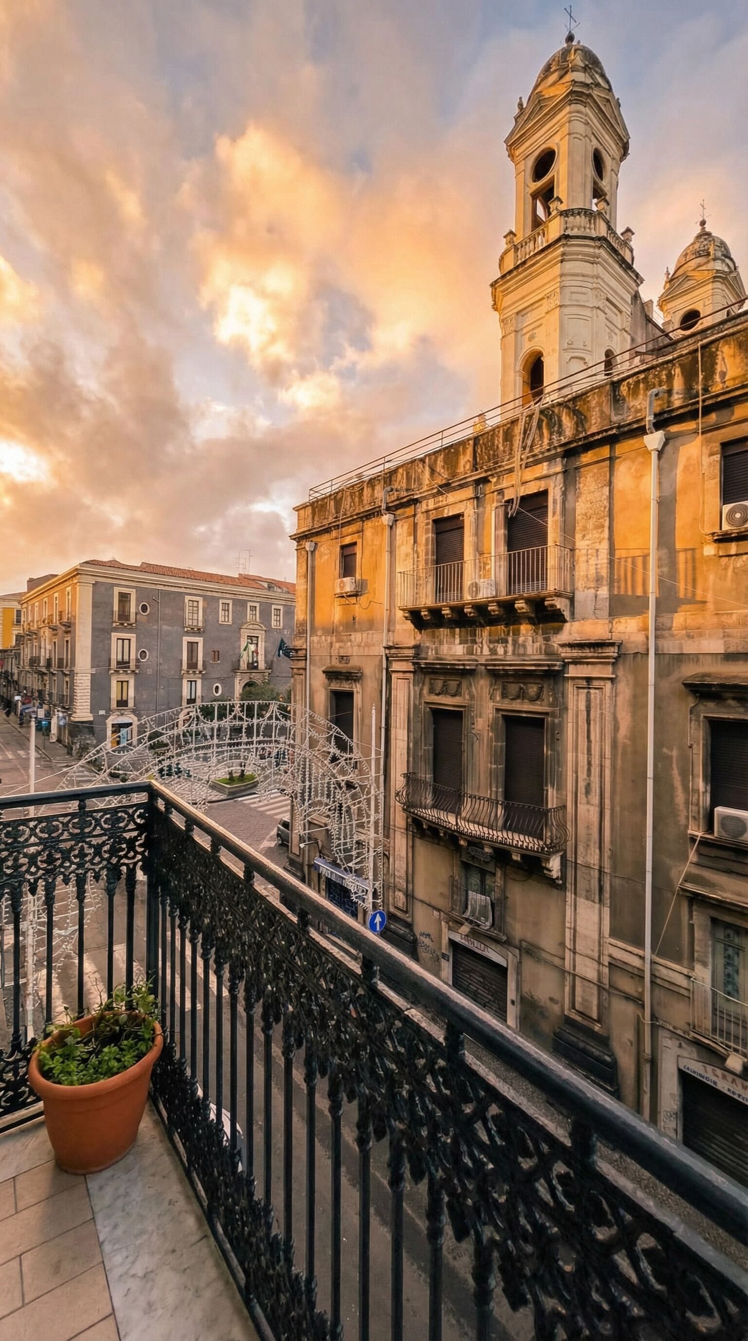 Uma varanda com um vaso de planta, com vista para um edifício histórico com uma torre de sino e uma cúpula, sob um céu nublado.