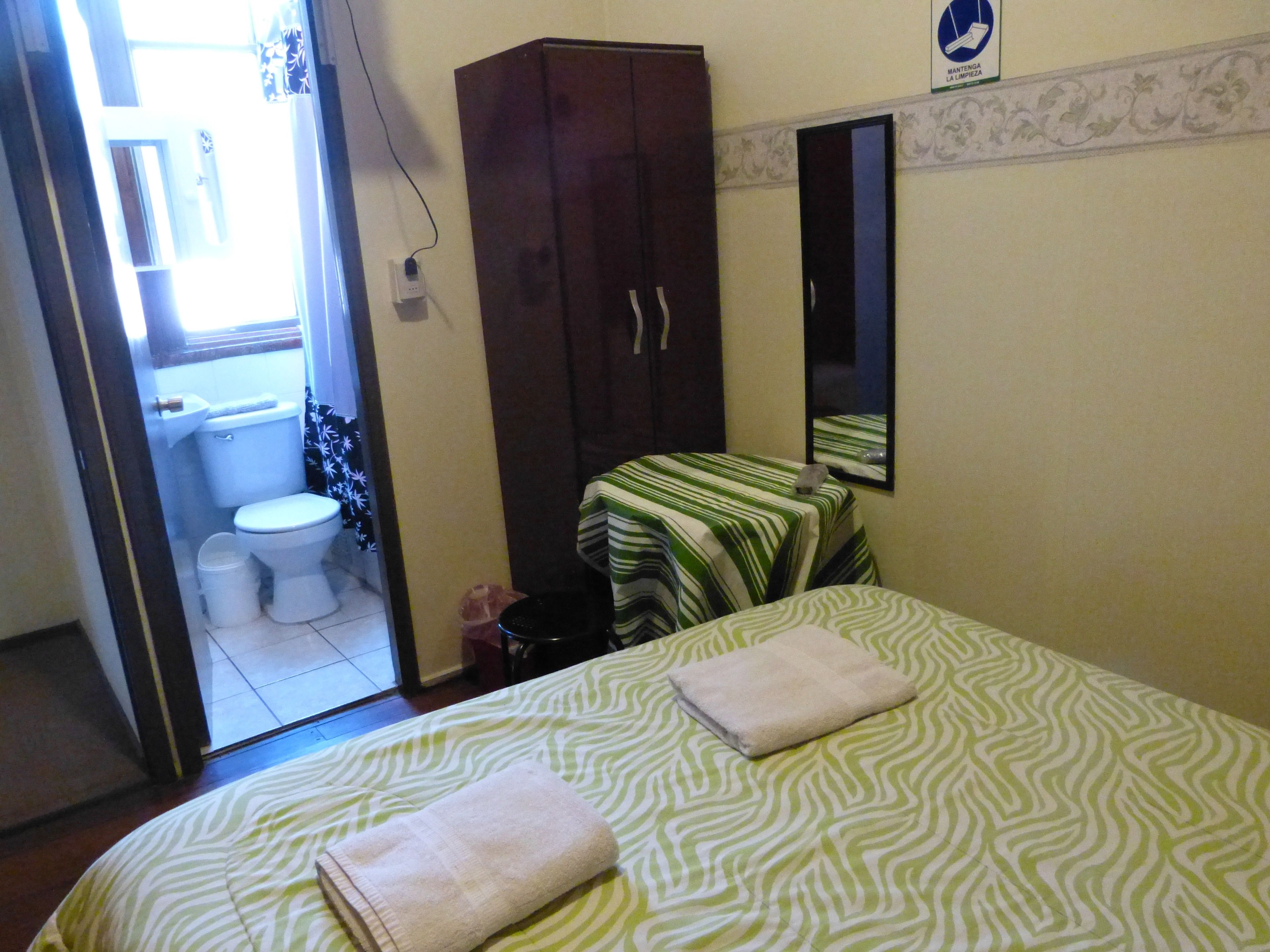 Quarto com cama, guarda-roupa, espelho e banheiro visível por uma porta aberta.