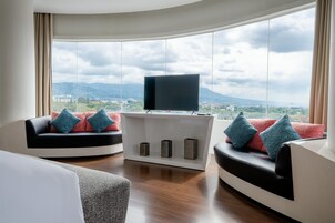 Quarto de hotel moderno com sofá curvo, televisão de tela plana e vista para a paisagem urbana através de grandes janelas.