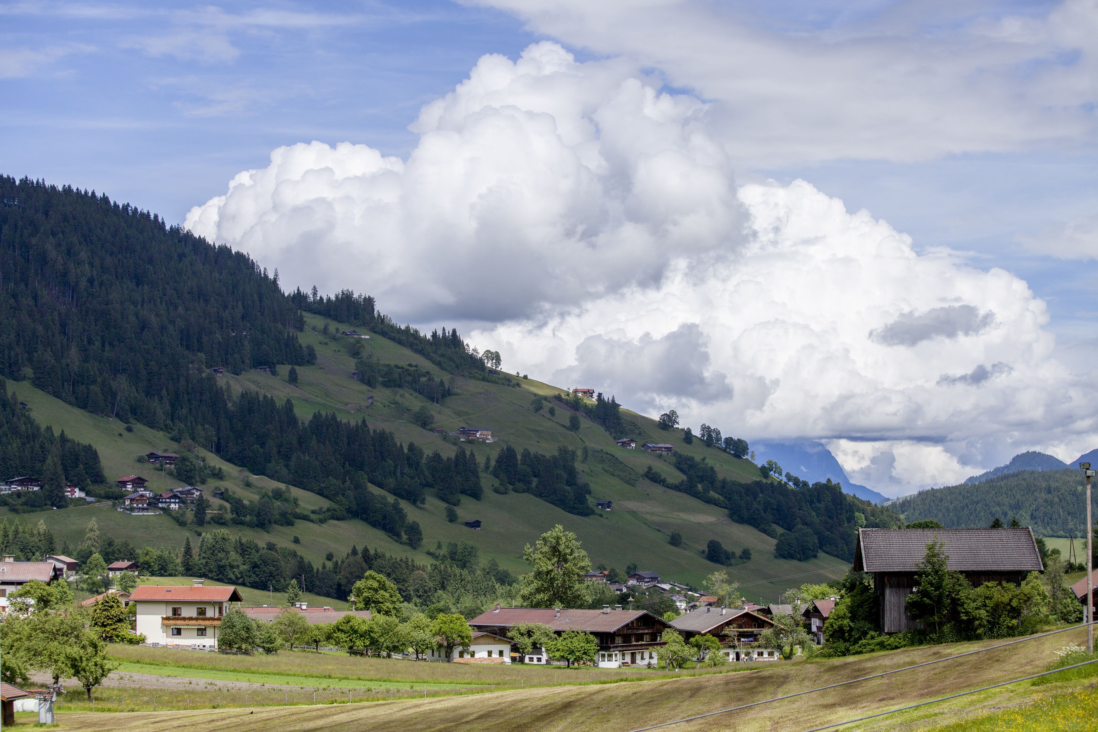 Uma vila rural com casas numa encosta, rodeada por campos verdes e montanhas.