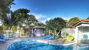 Piscina com cadeiras de descanso azuis, um pequeno gazebo e vegetação ao redor.