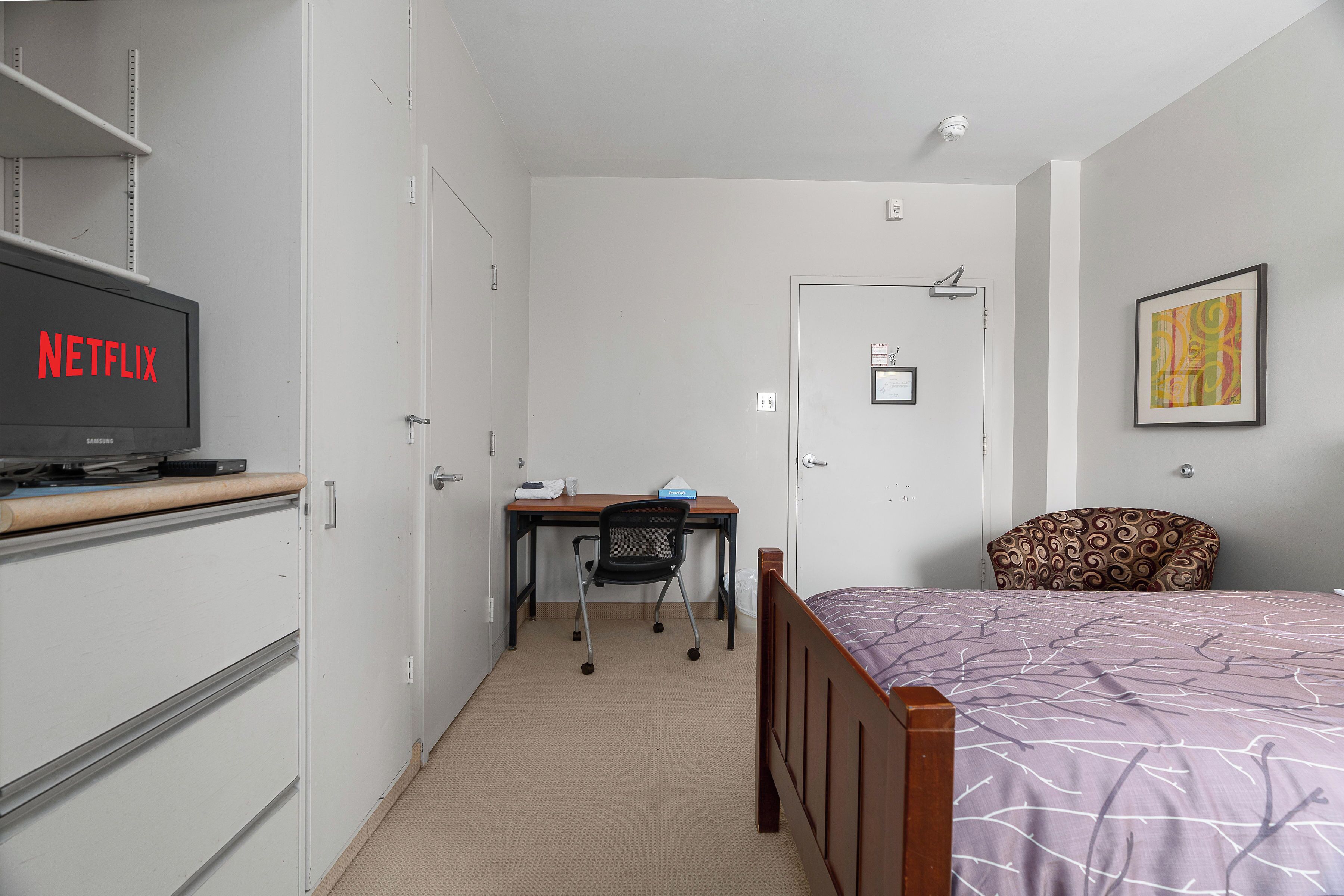 Quarto com uma cama, uma mesa com cadeira, uma televisão exibindo 'NETFLIX' e uma porta com moldura.