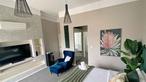 Quarto de hotel moderno com uma televisão de tela plana grande, uma poltrona azul confortável, um cobertor verde de tricô e uma planta grande.