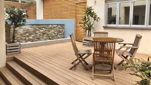 Um deck de madeira com uma mesa redonda e quatro cadeiras, uma parede de pedra e uma janela com persianas.