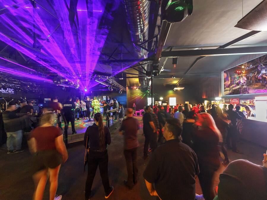 Um espaço interior lotado com palco e cabine de DJ, apresentando teto com vigas aparentes e luminárias.