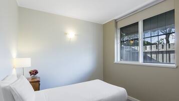 Quarto com uma cama, mesa de cabeceira com abajur e janela com vista para uma cerca e árvores.