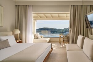 Quarto de hotel com uma cama grande, um sofá, uma pequena mesa com uma tigela de frutas e uma varanda com vista para a água e vegetação.