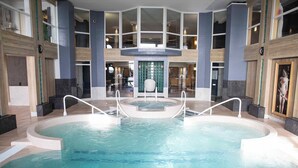Uma grande piscina interior com um ofurô central, rodeada por um átrio em vários níveis com áreas de estar e vista para o espaço interior.