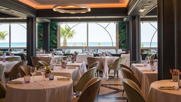 Restaurante com mesas redondas, cadeiras e vista para o mar.