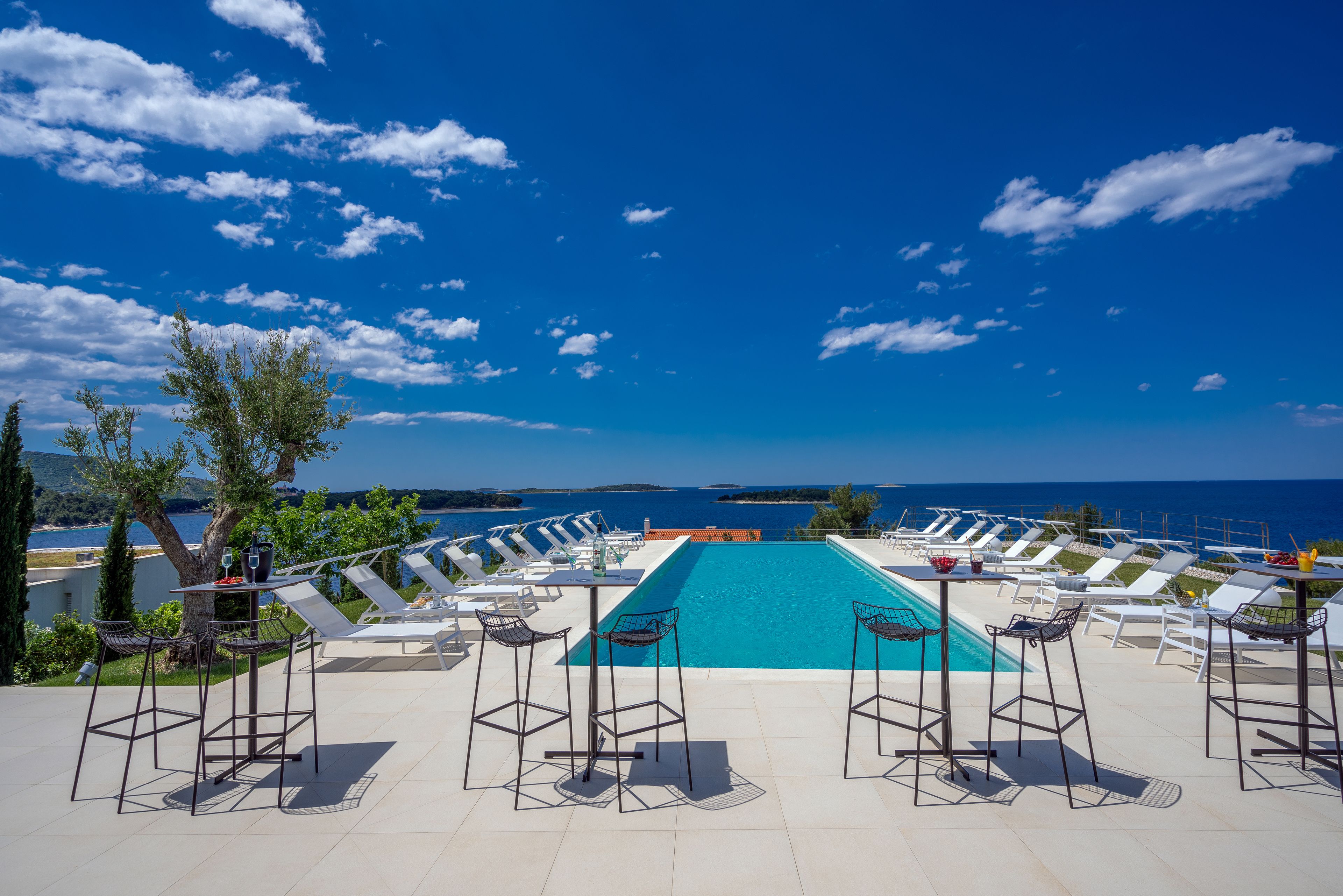 Área da piscina com espreguiçadeiras e uma mesa, com vista para um céu azul límpido e um corpo d'água.