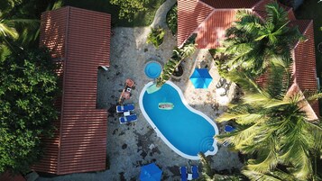 Vista aérea de um resort com piscina, cercada por cadeiras de descanso e guarda-sóis, palmeiras e edifícios com telhados vermelhos.