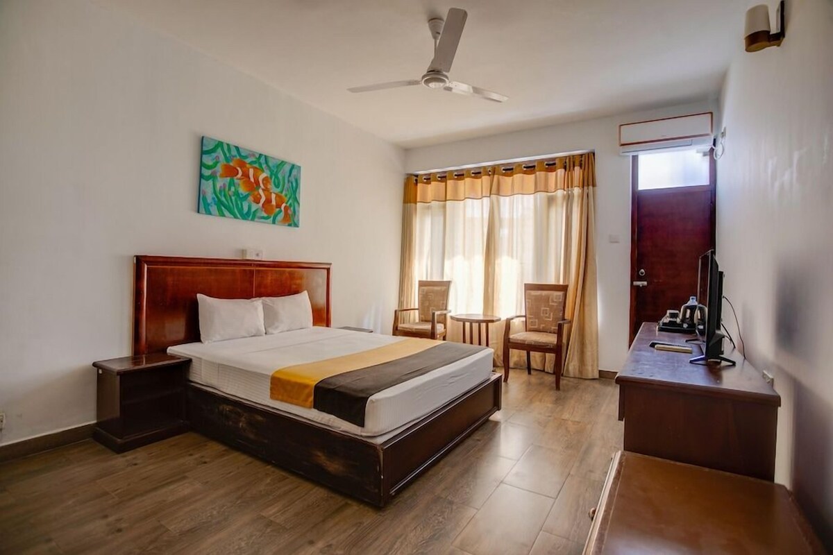 Quarto de hotel com cama, escrivaninha, cadeira, ventilador de teto e janela com cortinas.