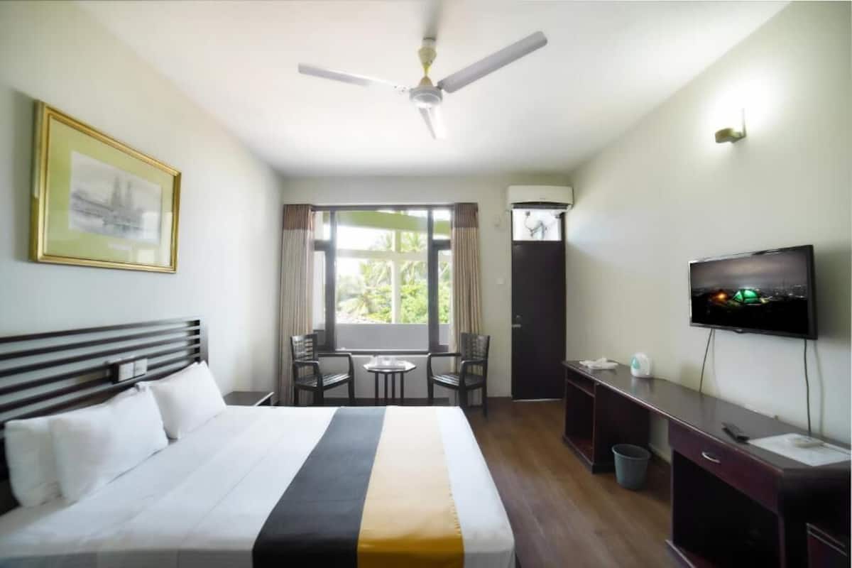 Quarto de hotel com cama, escrivaninha, televisão, ventoinha no teto e janela com cortinas.