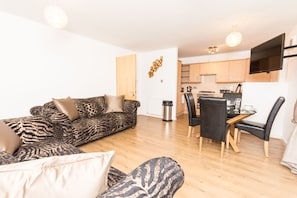 Uma sala moderna com sofá e poltronas, área de jantar com mesa e cadeiras, e cozinha com armários de madeira e uma televisão.