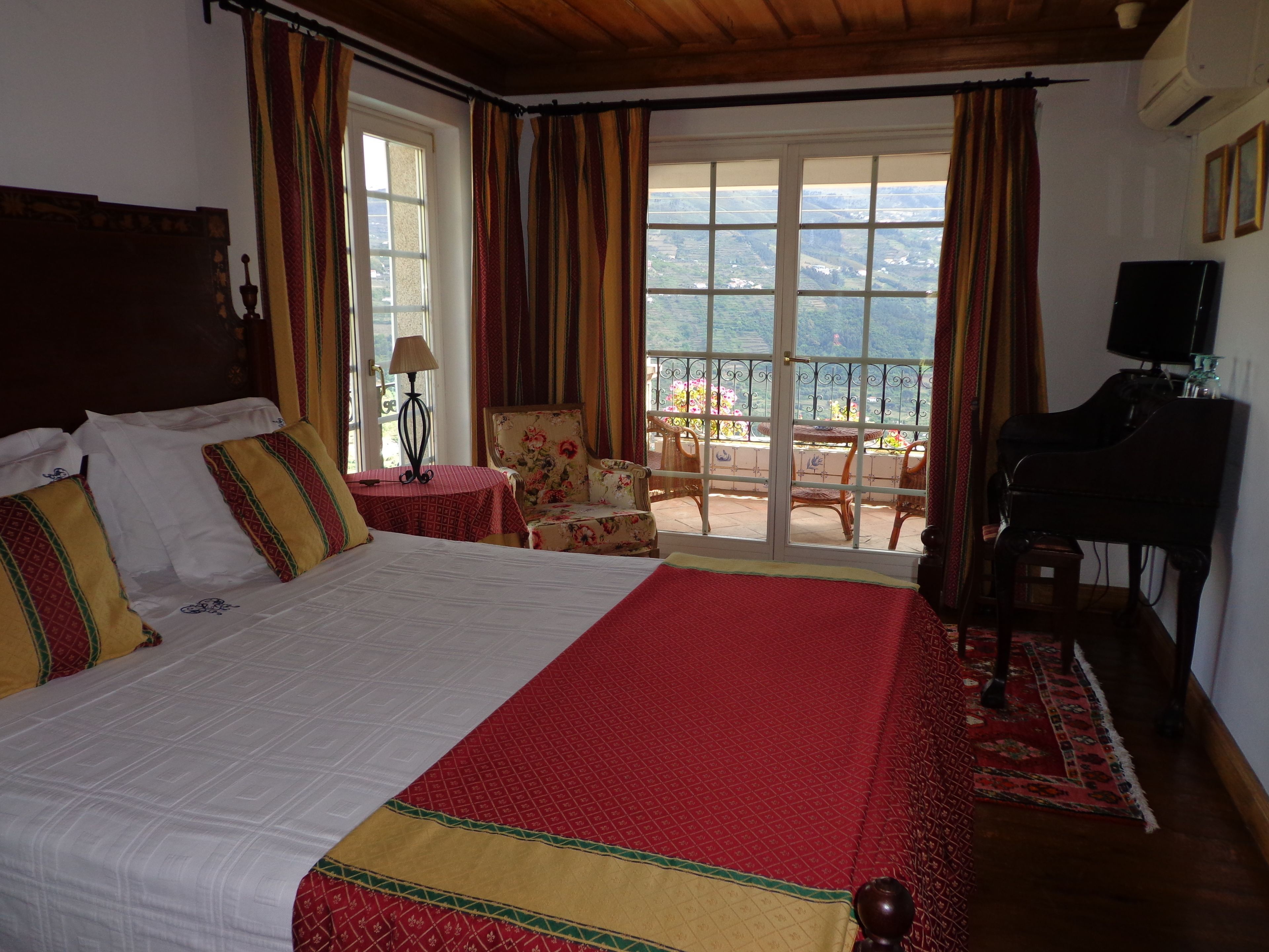 Quarto de hotel com uma cama grande, um piano, varanda com cadeiras e vista para o campo.