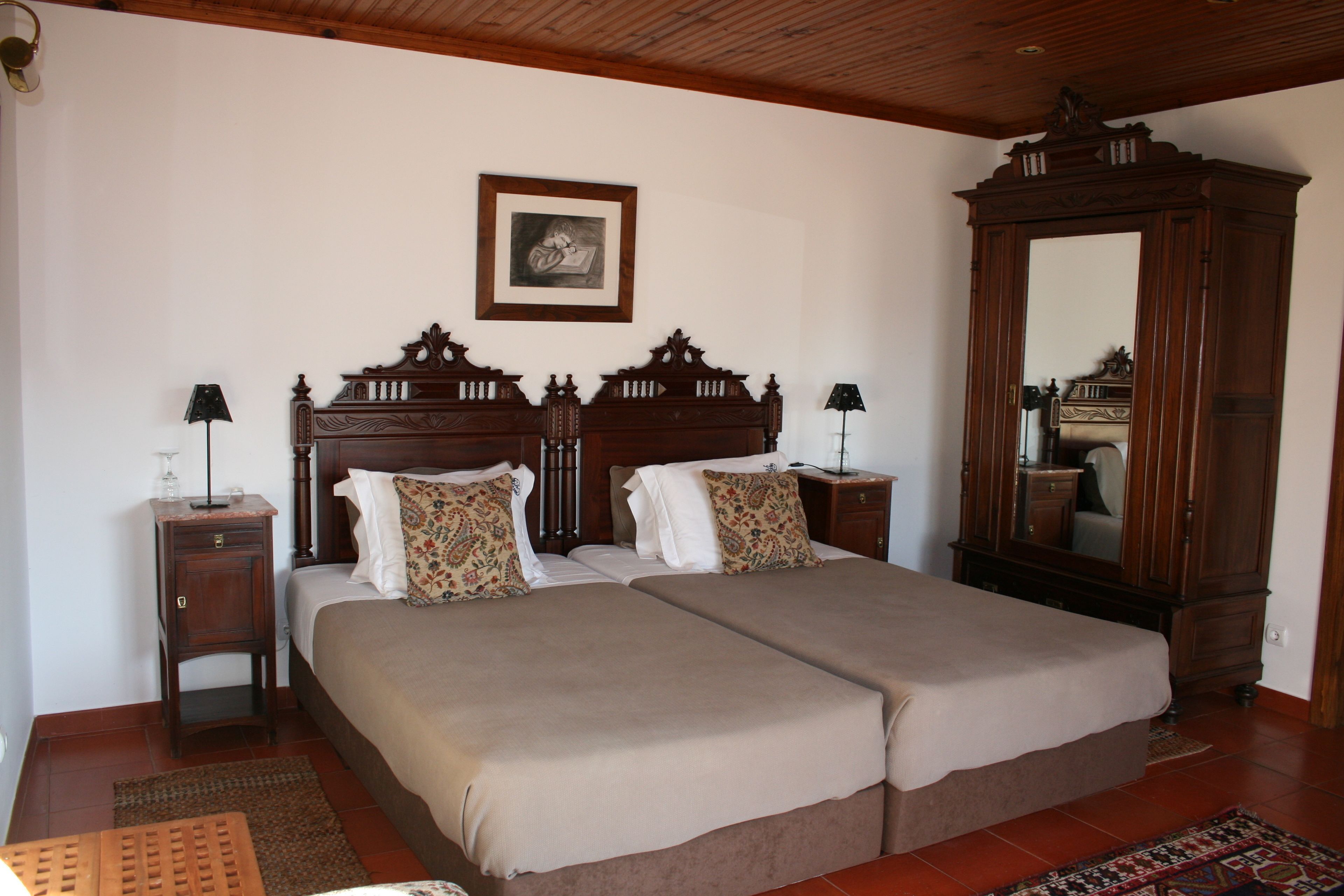 Uma cama de casal com cabeceira de madeira, duas mesas de cabeceira com abajures, um espelho grande e um quadro na parede.
