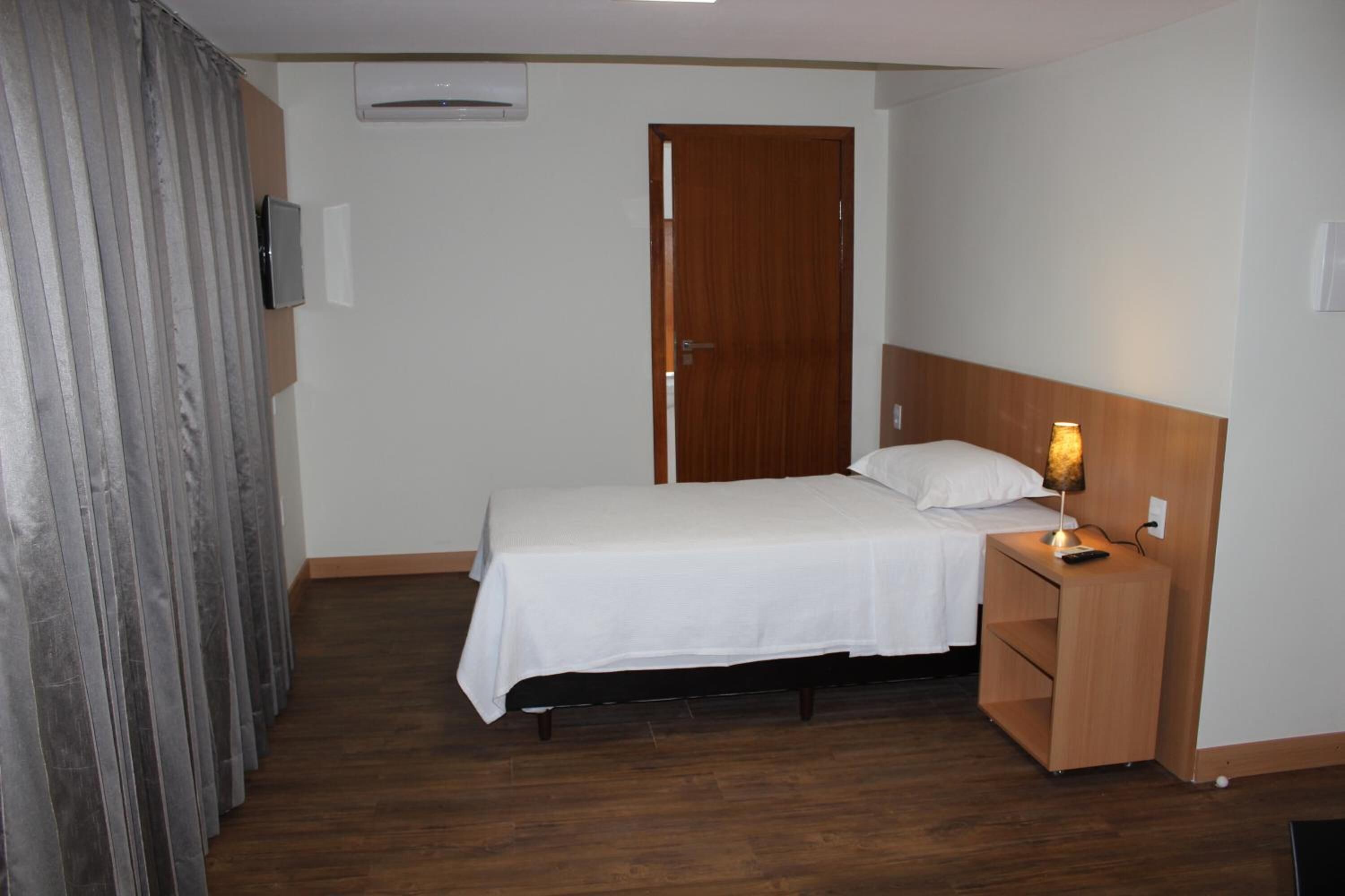 Uma cama de solteiro com cabeceira de madeira, uma mesa de cabeceira com abajur e uma cortina à esquerda.