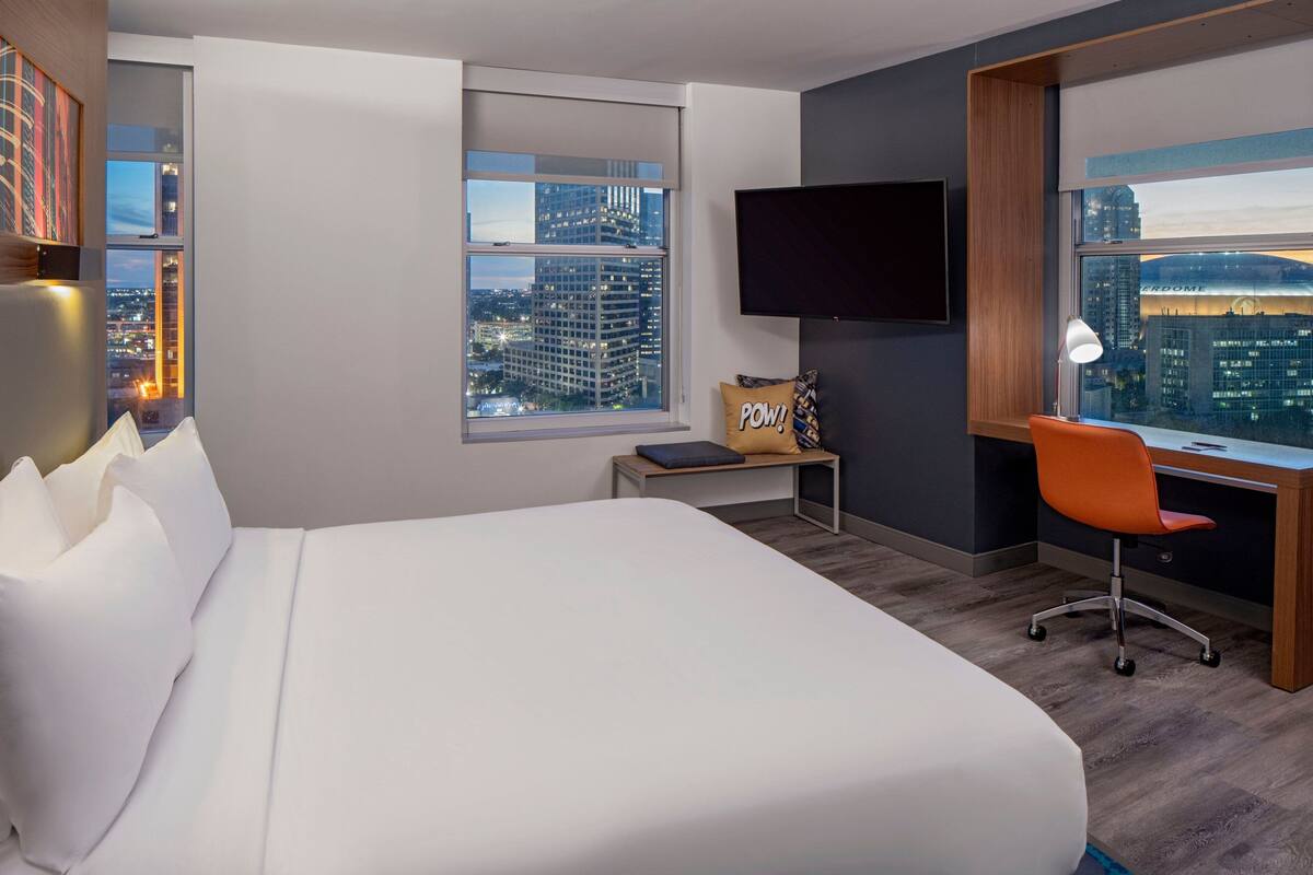 Quarto de hotel moderno com uma cama grande, uma mesa com uma cadeira laranja, uma TV montada na parede e vista para a cidade por grandes janelas.