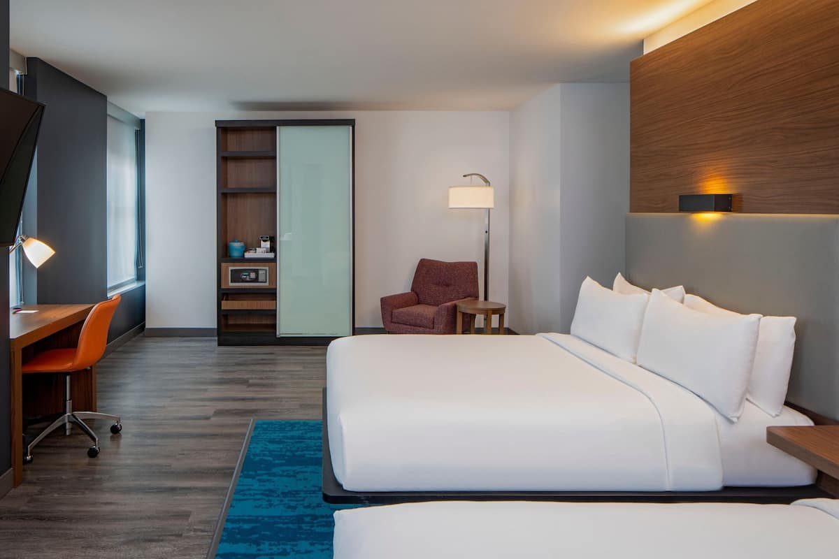 Quarto de hotel com uma cama grande, uma escrivaninha com cadeira, uma mesinha, uma luminária e um guarda-roupa.