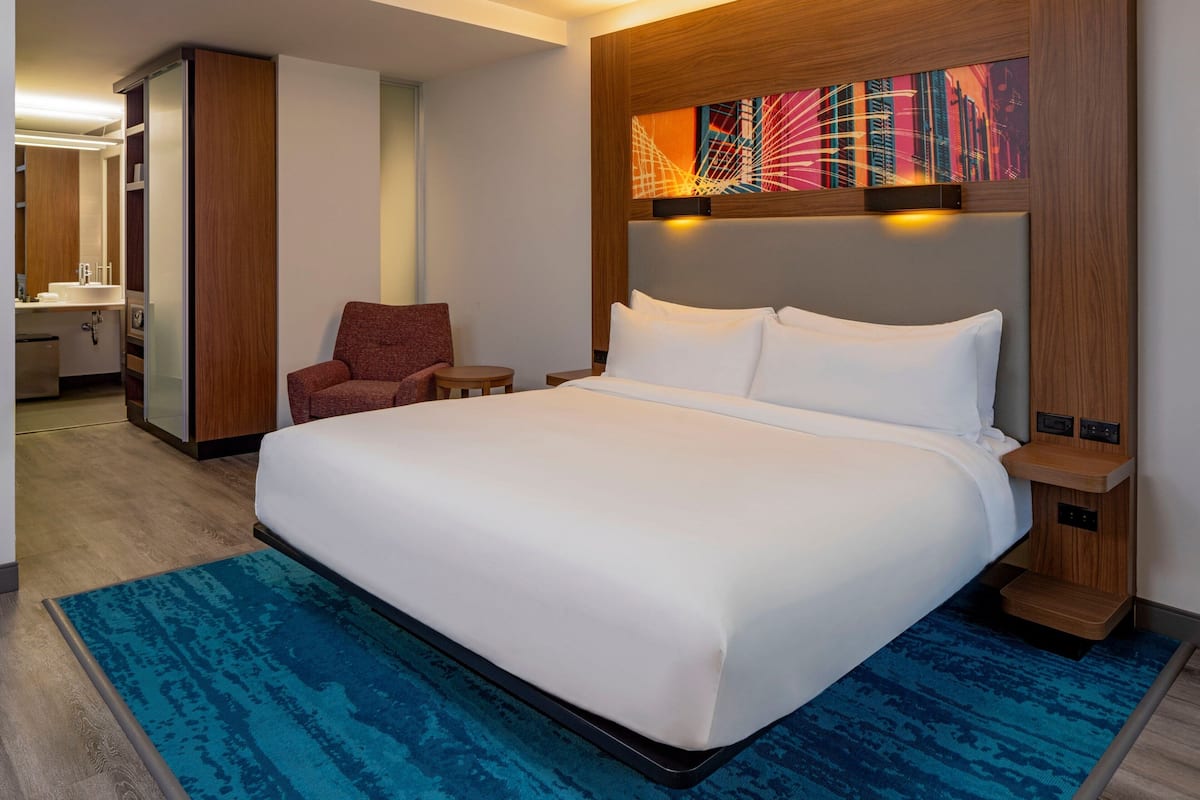 Quarto de hotel com uma cama grande, uma cadeira, uma mesinha e uma mesa de cabeceira.