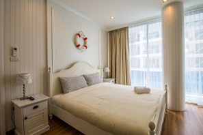 Quarto de hotel com uma cama grande, uma mesa de cabeceira com abajur, um salva-vidas fixado na parede e uma janela com cortinas.