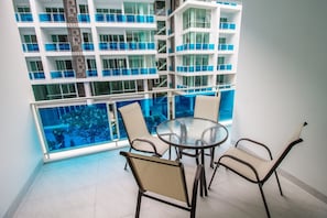 Varanda com mesa e cadeiras de vidro, com vista para uma piscina e um edifício com várias varandas.