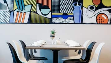 Uma área de refeições com uma mesa posta para quatro pessoas, rodeada por cadeiras modernas, e uma pintura abstrata colorida na parede.