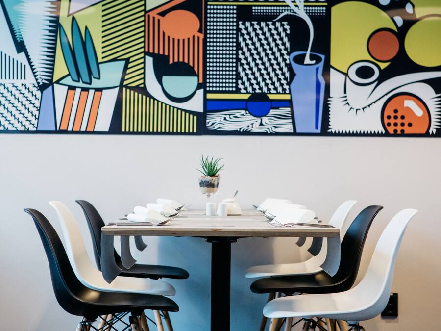 Uma área de refeições com uma mesa posta para quatro pessoas, rodeada por cadeiras modernas, e uma pintura abstrata colorida na parede.