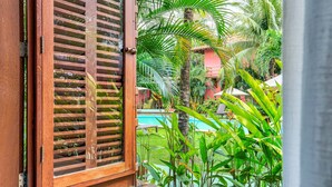 Uma porta de madeira com ripas horizontais, um deck de madeira e vista para uma piscina cercada por vegetação exuberante e palmeiras.
