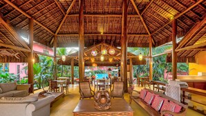 Um espaço amplo e coberto, com teto de palha, móveis de madeira e uma piscina visível através das paredes de vidro.