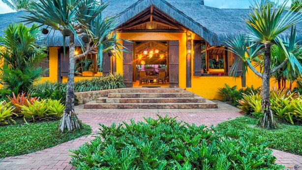 Um resort tropical com um prédio de telhado de palha, entrada pavimentada e vegetação exuberante.