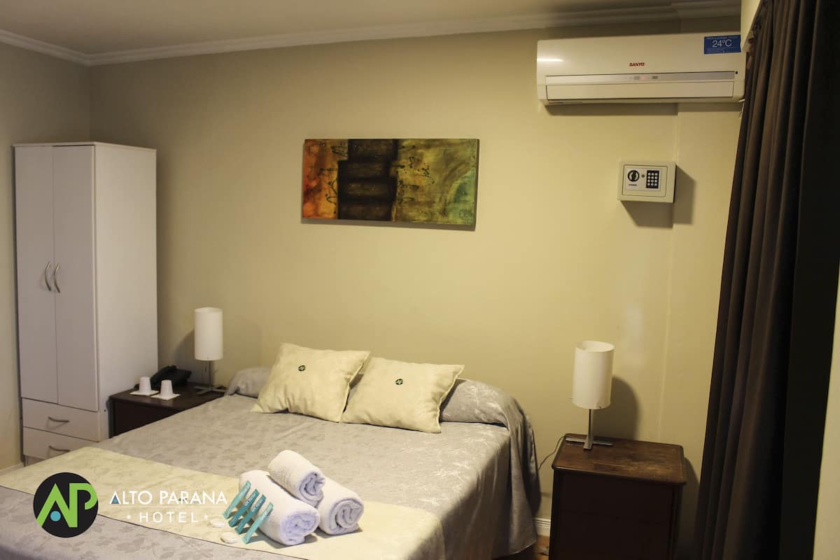 Quarto de hotel com cama, guarda-roupa, um quadro na parede, ar condicionado e painel de controle.