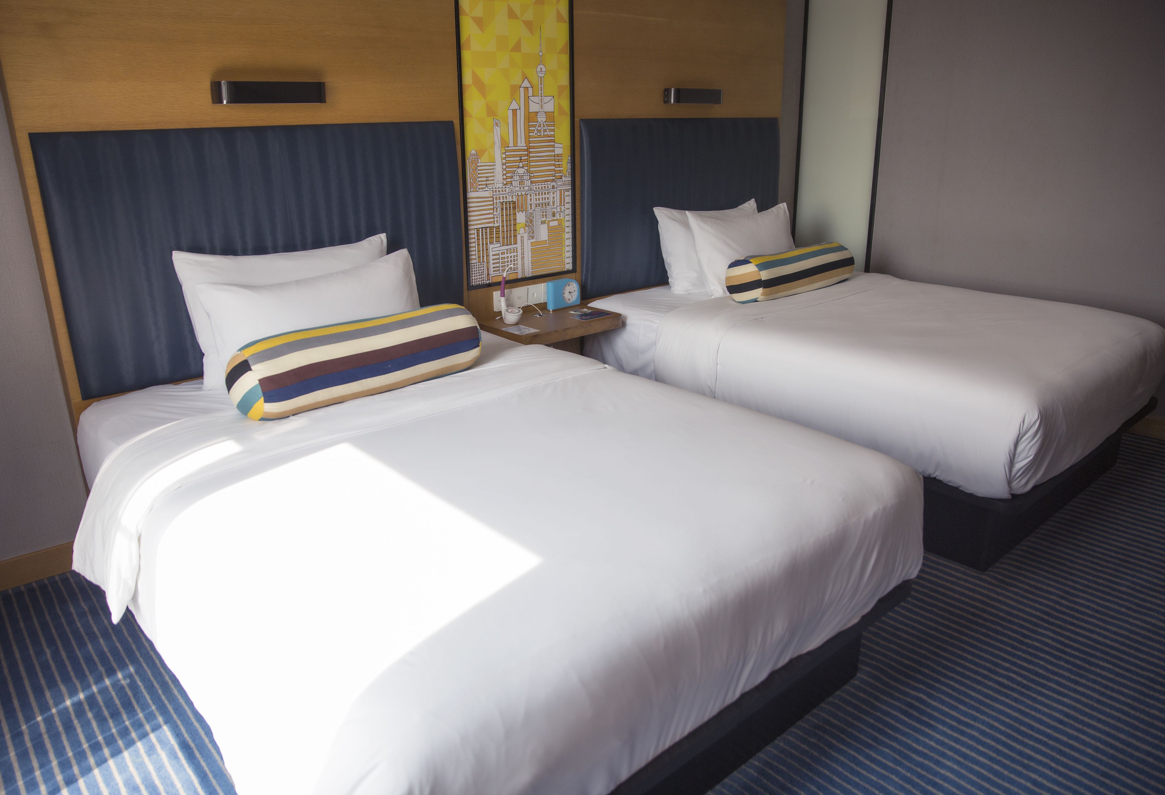 Duas camas de solteiro com roupa de cama branca, uma cabeceira de madeira, uma obra de arte com tema urbano e uma pequena mesa com um relógio e uma xícara.