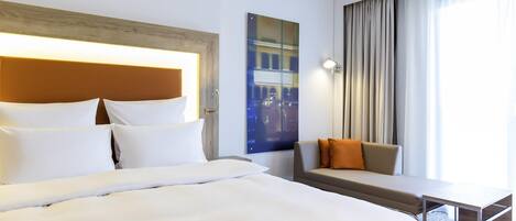 Quarto de hotel com uma cama grande, um sofá, uma mesinha e uma janela ampla com cortinas.