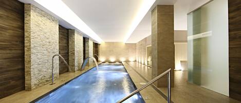 Uma área interna de piscina moderna com paredes de pedra, piscina azul e uma divisória de vidro de um lado.