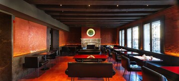 Interior de restaurante com paredes de tijolo, vigas de madeira e lareira central. Mesas e cadeiras dispostas para refeições.