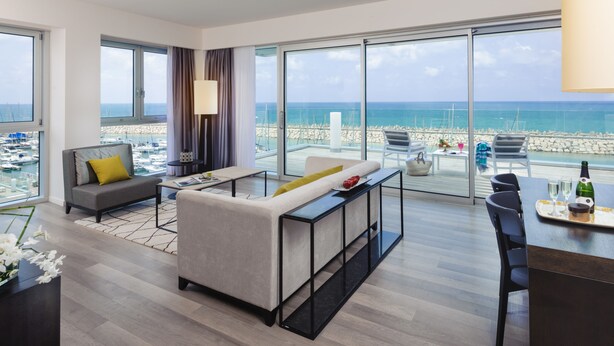 Quarto de hotel moderno com varanda ampla, sofá, área de jantar e vista para a praia e o oceano.
