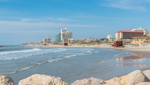 Uma praia com pedras em primeiro plano, um hotel com vários andares e um edifício com telhado vermelho ao fundo.
