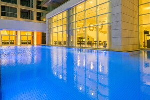 Piscina interior com água azul cristalina, rodeada por um edifício moderno com grandes janelas e uma escadaria que leva a um andar superior.