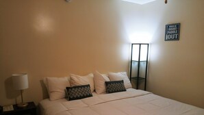 Cama bem arrumada com roupa de cama branca e duas almofadas decorativas, abajur e placa na parede.