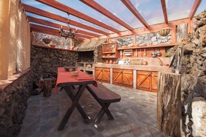 Uma cozinha rústica ao ar livre, com parede de pedra, vigas de madeira e mesa de madeira com banco.