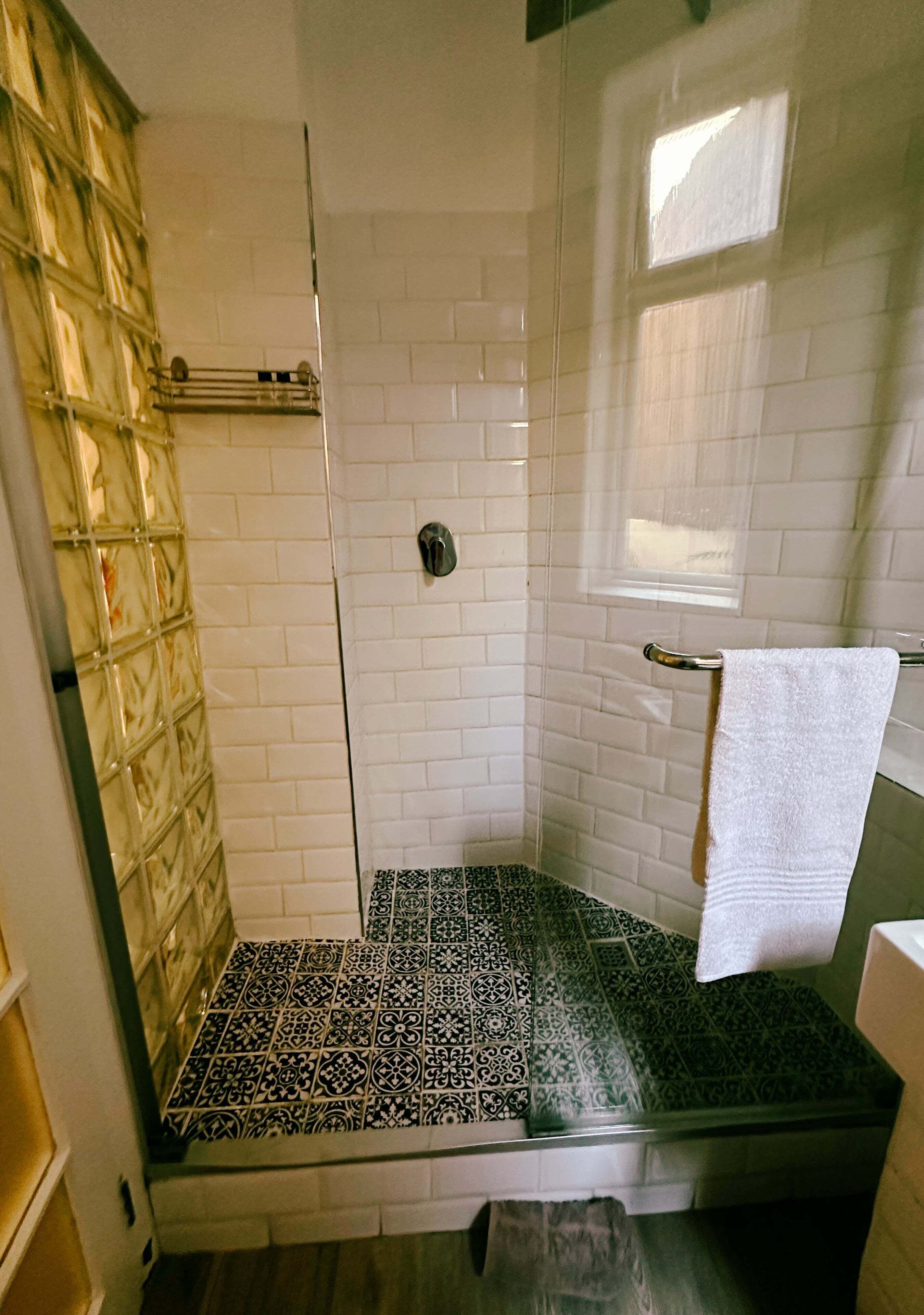 Chuveiro com porta de vidro, paredes com azulejos brancos e chão estampado.