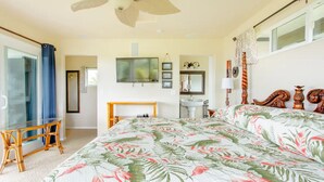 Quarto com cama grande, escrivaninha, ventilador de teto, janela com cortinas e banheiro com pia e espelho.