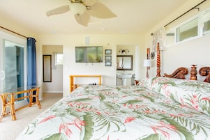 Quarto com cama grande, escrivaninha, ventilador de teto, janela com cortinas e banheiro com pia e espelho.