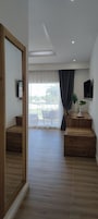 Quarto moderno com um banco de madeira, uma pequena mesa com um vaso, e uma TV fixada na parede. Há um espelho grande à esquerda e vista para uma área externa com um banco e uma mesa.