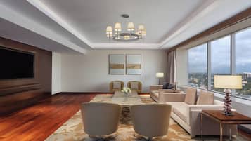 Quarto de hotel moderno com uma televisão de tela plana grande, área de jantar com mesa e cadeiras, um conjunto de sofás e uma grande janela com vista para a cidade.