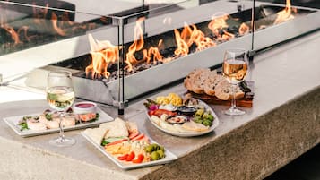 Uma mesa de jantar com diversos pratos e bebidas, incluindo uma taça de vinho, e uma lareira ao fundo.