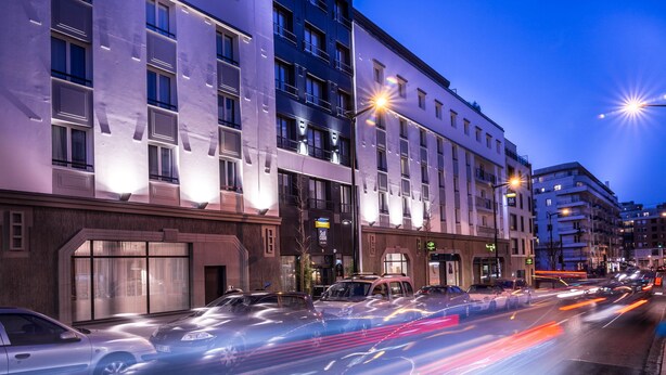 Um hotel com vários andares, fachada moderna, janelas iluminadas e uma rua movimentada com rastros de luz deixados por veículos em movimento.